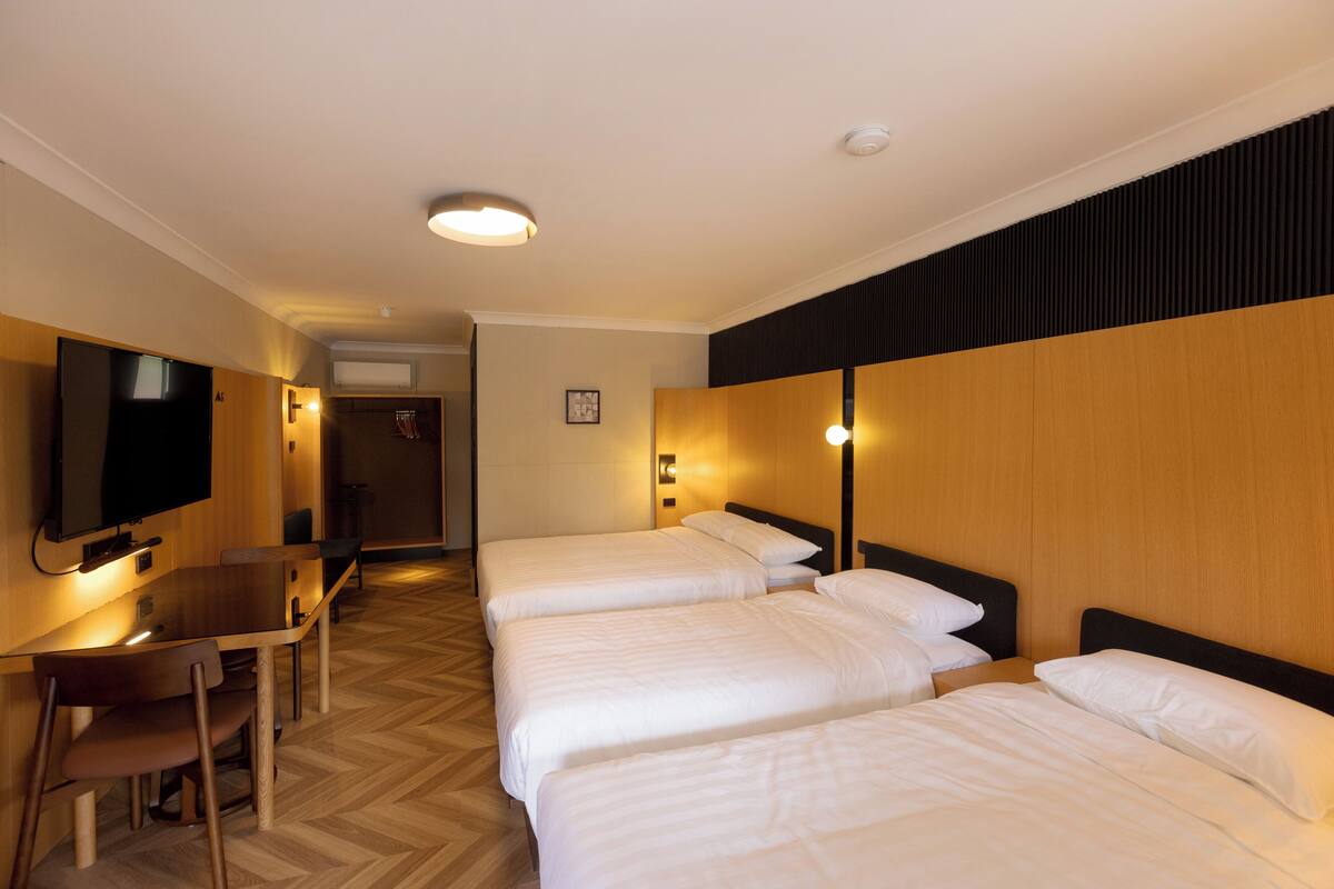 Quarto de hotel com três camas, uma escrivaninha com cadeira, televisão e luminária fixada na parede.
