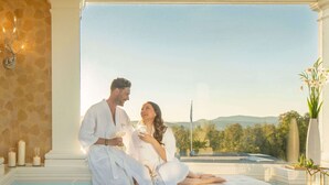 Um casal sentado num sofá branco numa sala com uma janela grande que dá para uma paisagem deslumbrante.
