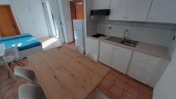 Cozinha compacta com mesa de madeira, cadeiras brancas, pia e geladeira.