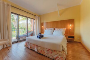 Quarto de hotel com uma cama grande, cabeceira de madeira, varanda com uma cadeira e vista para palmeiras.