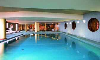 Uma grande piscina interna com superfície de água azul cristalina, rodeada por espreguiçadeiras e um design moderno com janelas circulares e azulejos decorativos.