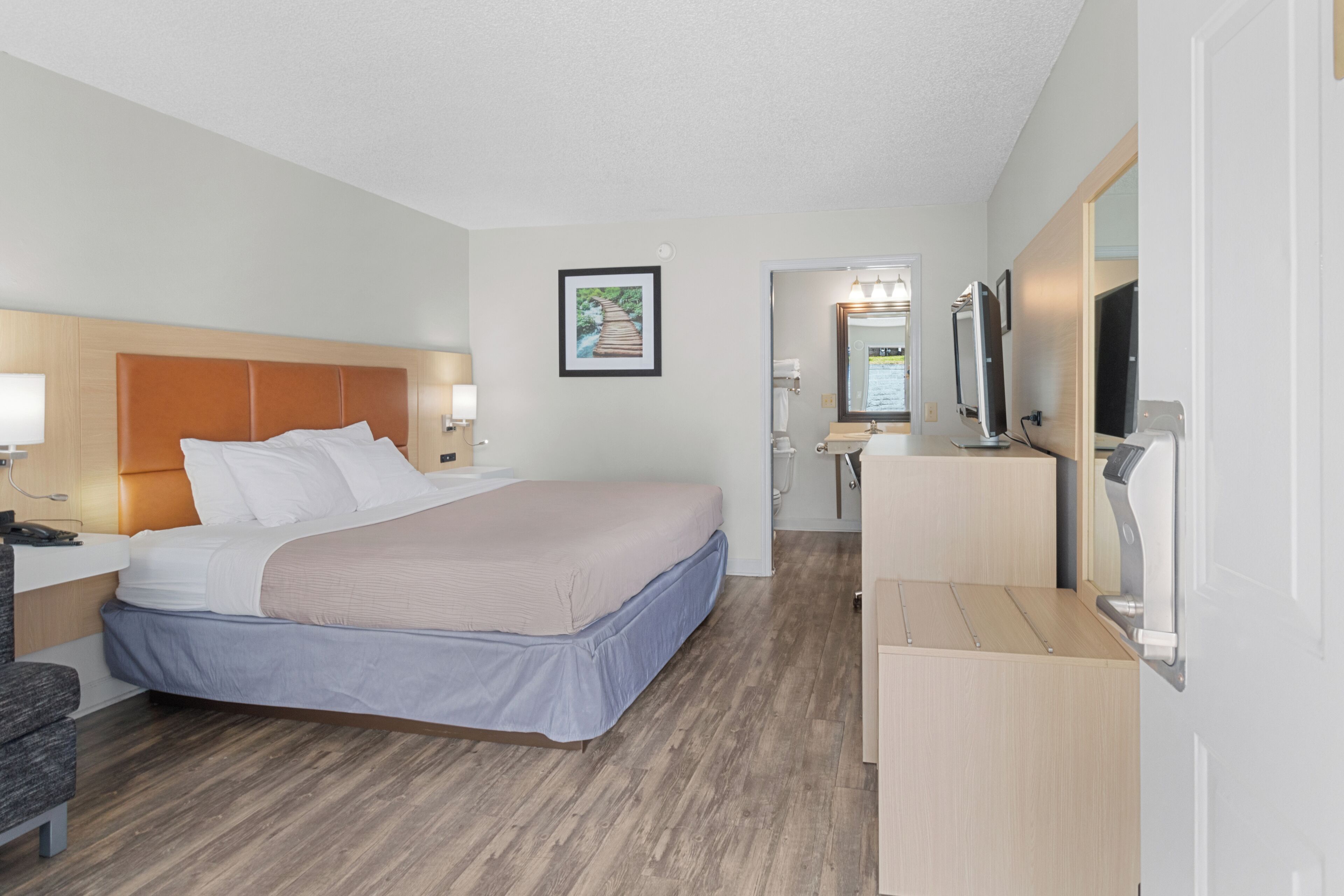 Quarto de hotel com cama, mesa de cabeceira, luminária e escrivaninha com cadeira.