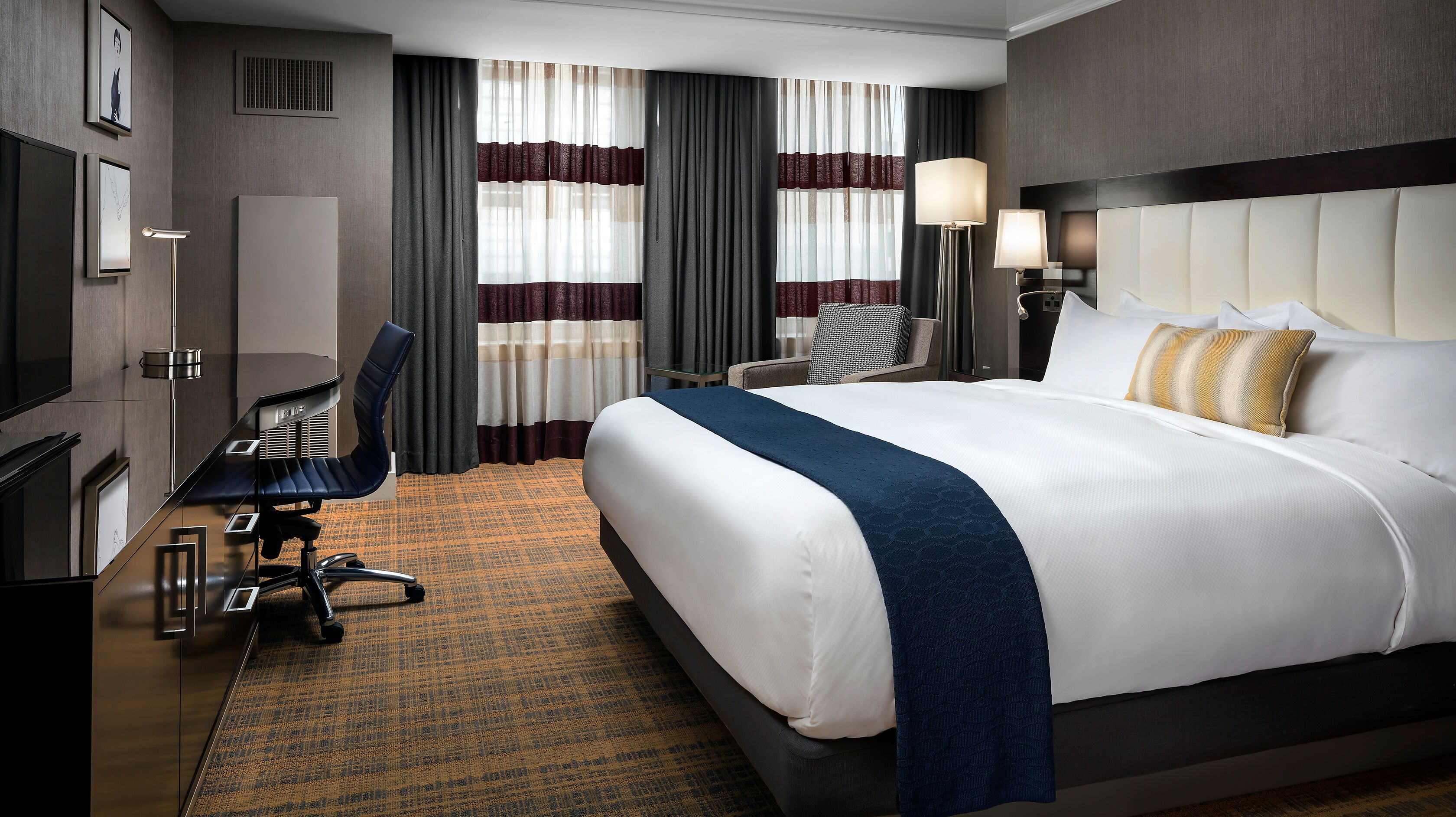 Quarto de hotel com uma cama grande, uma escrivaninha com cadeira, luminária e televisão.