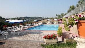 Uma área de piscina com espreguiçadeiras e guarda-sóis, cercada por canteiros de flores e um muro de pedra.