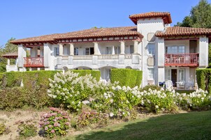 Um edifício grande de dois andares com telhado de telhas vermelhas, várias varandas e um jardim bem cuidado com diversas plantas floridas.