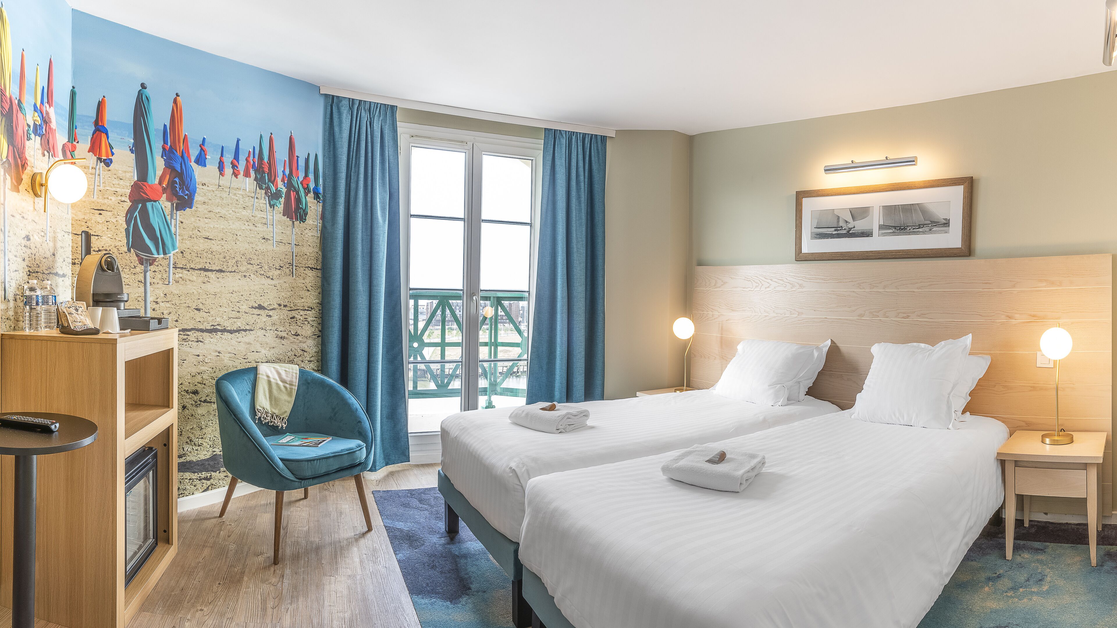Quarto de hotel com duas camas, uma escrivaninha, uma cadeira e vista para a praia pela janela.