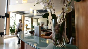 Um hall de hotel moderno com uma recepção de vidro, um vaso com orquídeas brancas e uma pessoa sentada em uma mesa ao fundo.