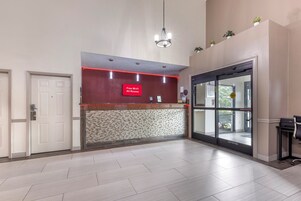 Uma área de recepção de hotel com uma placa vermelha indicando Wi-Fi gratuito, uma luminária moderna e uma porta de vidro que dá para o exterior.