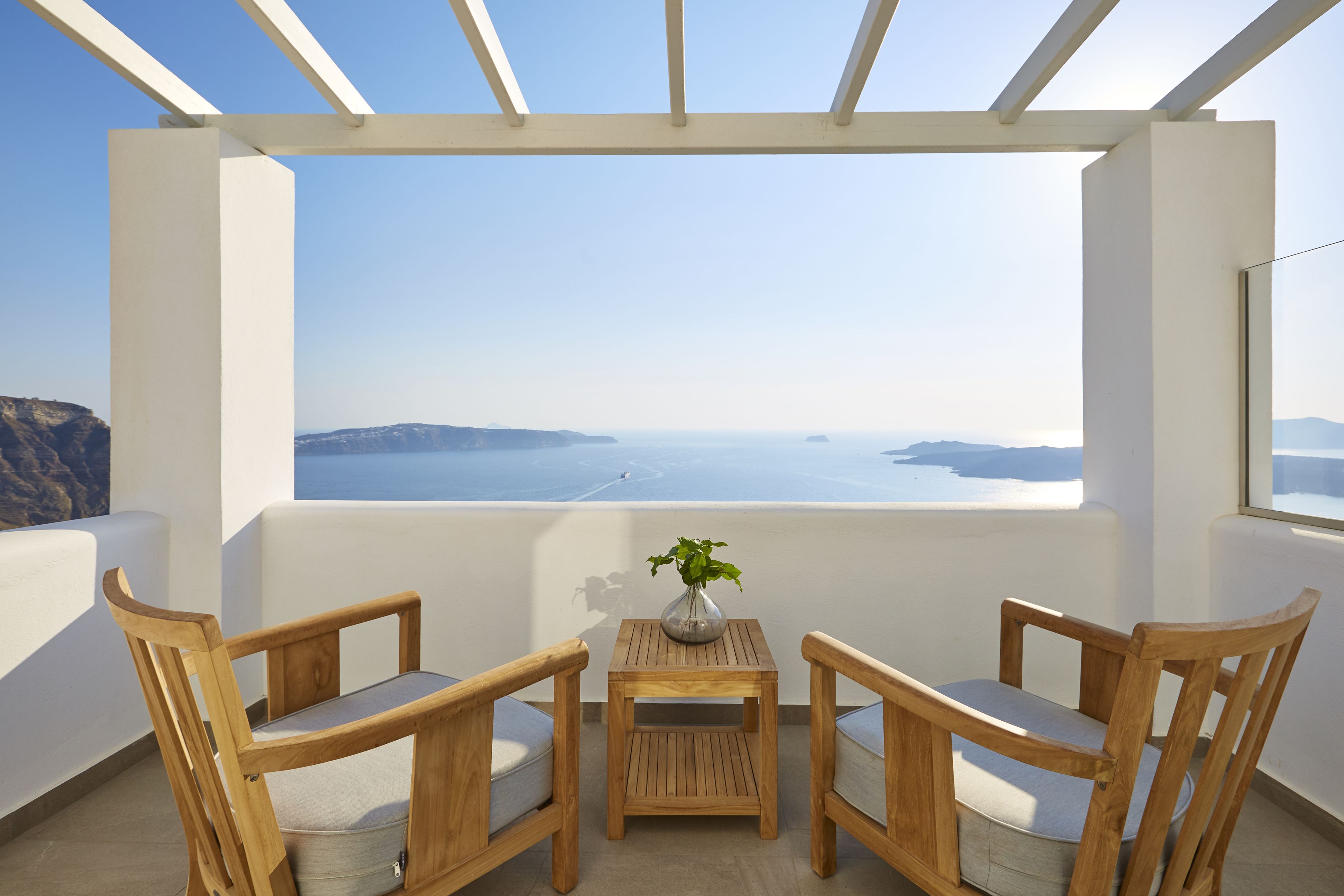 Varanda com mesa de madeira e duas cadeiras, vaso com planta e vista para o mar e terra ao longe.