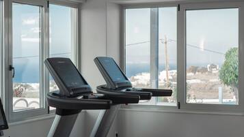 Uma academia com esteiras e bicicletas ergométricas, grandes janelas com vista para o mar e uma luminária no teto.