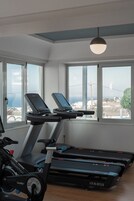 Uma academia com esteiras e bicicletas ergométricas, grandes janelas com vista para o mar e uma luminária no teto.