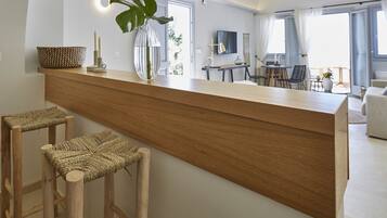 Uma cozinha moderna com bancada de madeira, dois taboretes trançados, uma planta grande e uma área de jantar com mesa e cadeiras.