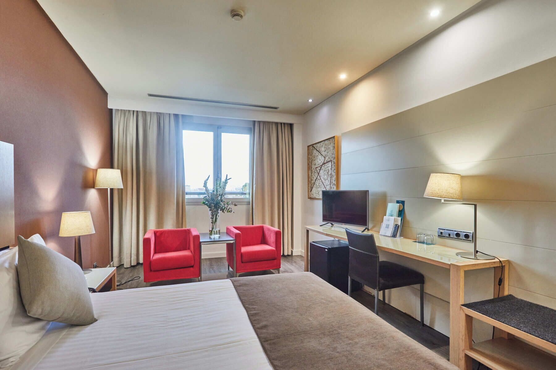Quarto de hotel com uma cama, duas cadeiras vermelhas, uma escrivaninha com um computador, uma televisão e uma janela com cortinas.