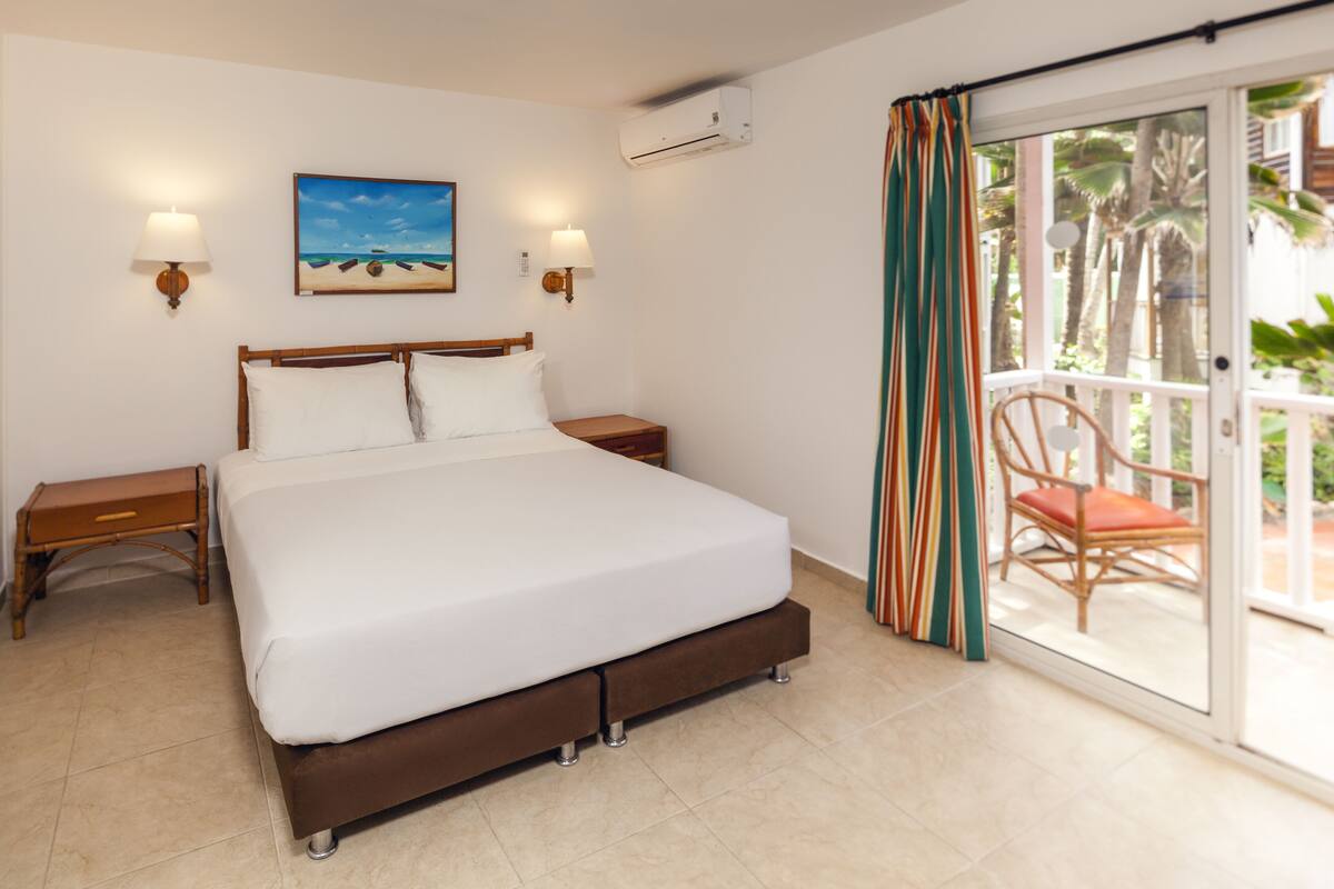 Quarto com cama, luminárias de cabeceira, um quadro, ar-condicionado e vista para uma varanda com uma cadeira.