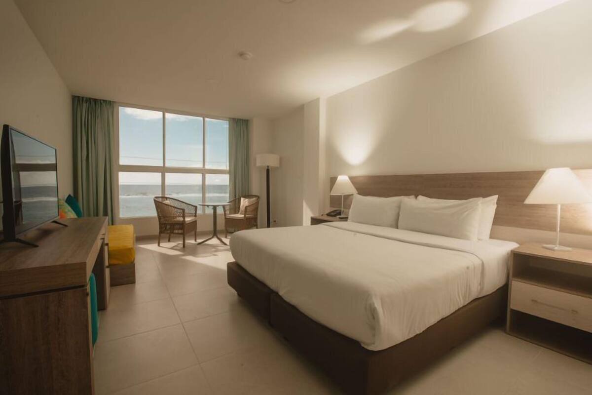 Quarto de hotel com uma cama grande, uma mesa com televisão, uma cadeira, uma mesa e vista para o oceano.