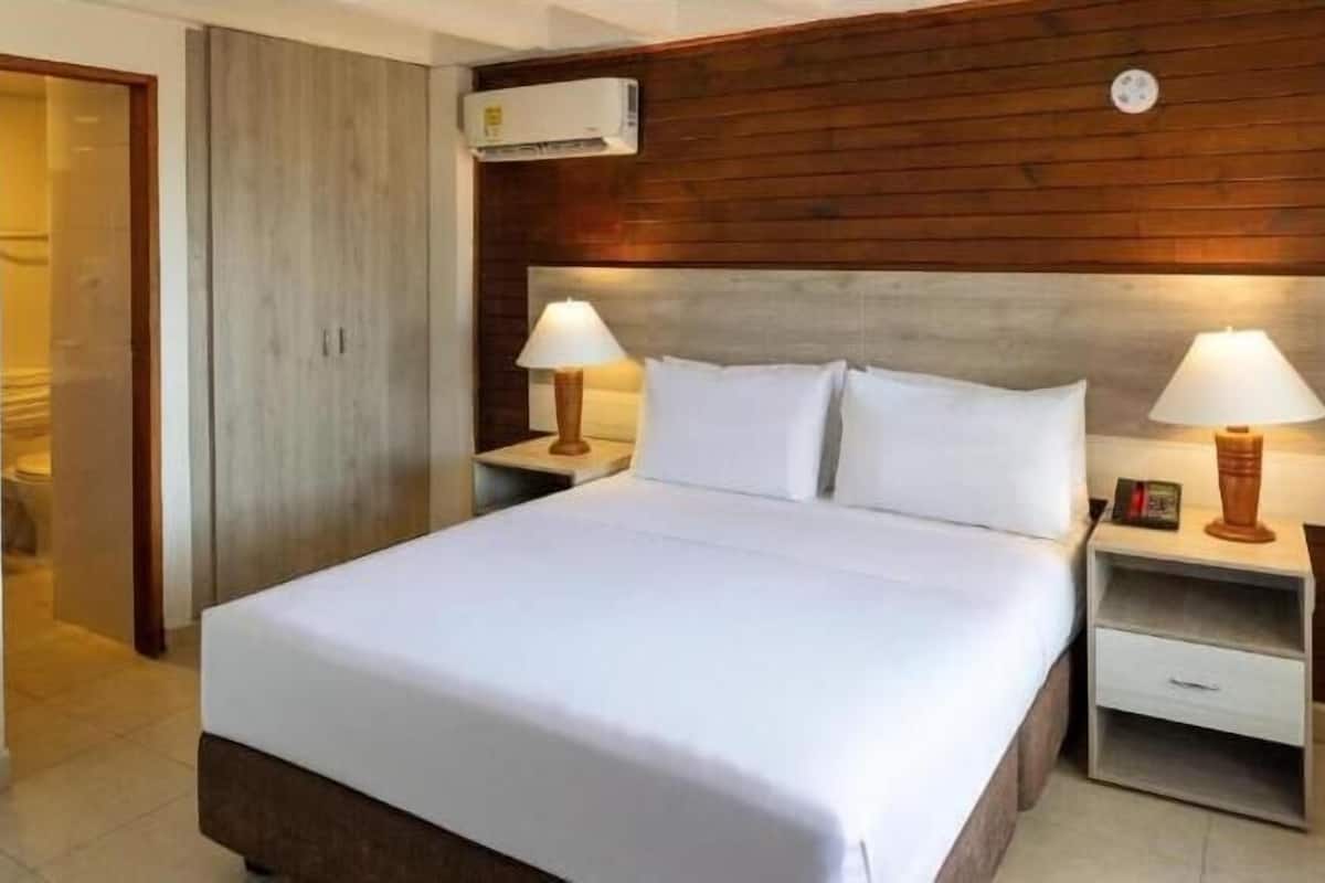 Quarto de hotel com cabeceira de madeira, uma cama grande com lençóis brancos, duas mesas de cabeceira com abajures e um guarda-roupa.