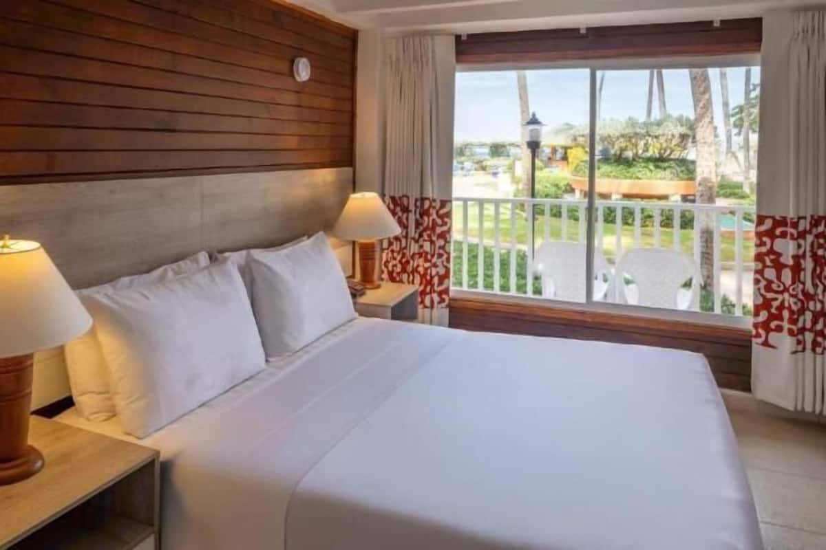 Quarto de hotel com cabeceira de madeira, uma cama grande com roupa de cama branca, duas luminárias de cabeceira e uma varanda com vista para o verde e uma piscina.