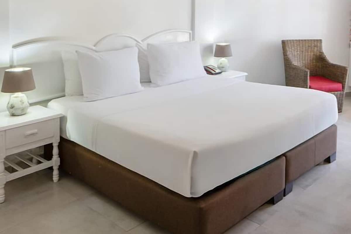 Cama bem arrumada com roupa de cama branca, mesa de cabeceira com abajur e cadeira com almofada vermelha.