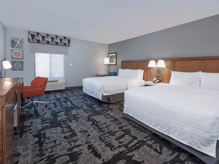 Quarto de hotel com duas camas, uma escrivaninha, uma cadeira e uma janela grande.