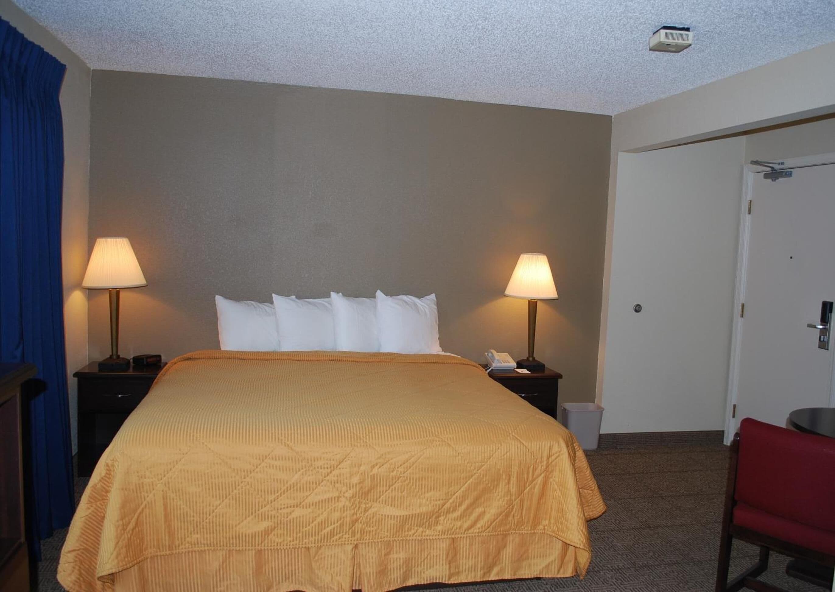 Quarto de hotel com uma cama grande, duas mesas de cabeceira com abajures, uma cadeira vermelha e uma porta.