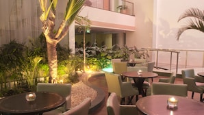 Restaurante com mesas e cadeiras redondas, cercado por plantas e uma palmeira.
