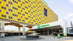 Um hotel Holiday Inn com fachada distinta em amarelo e preto, entrada coberta e um pátio central com uma fonte.