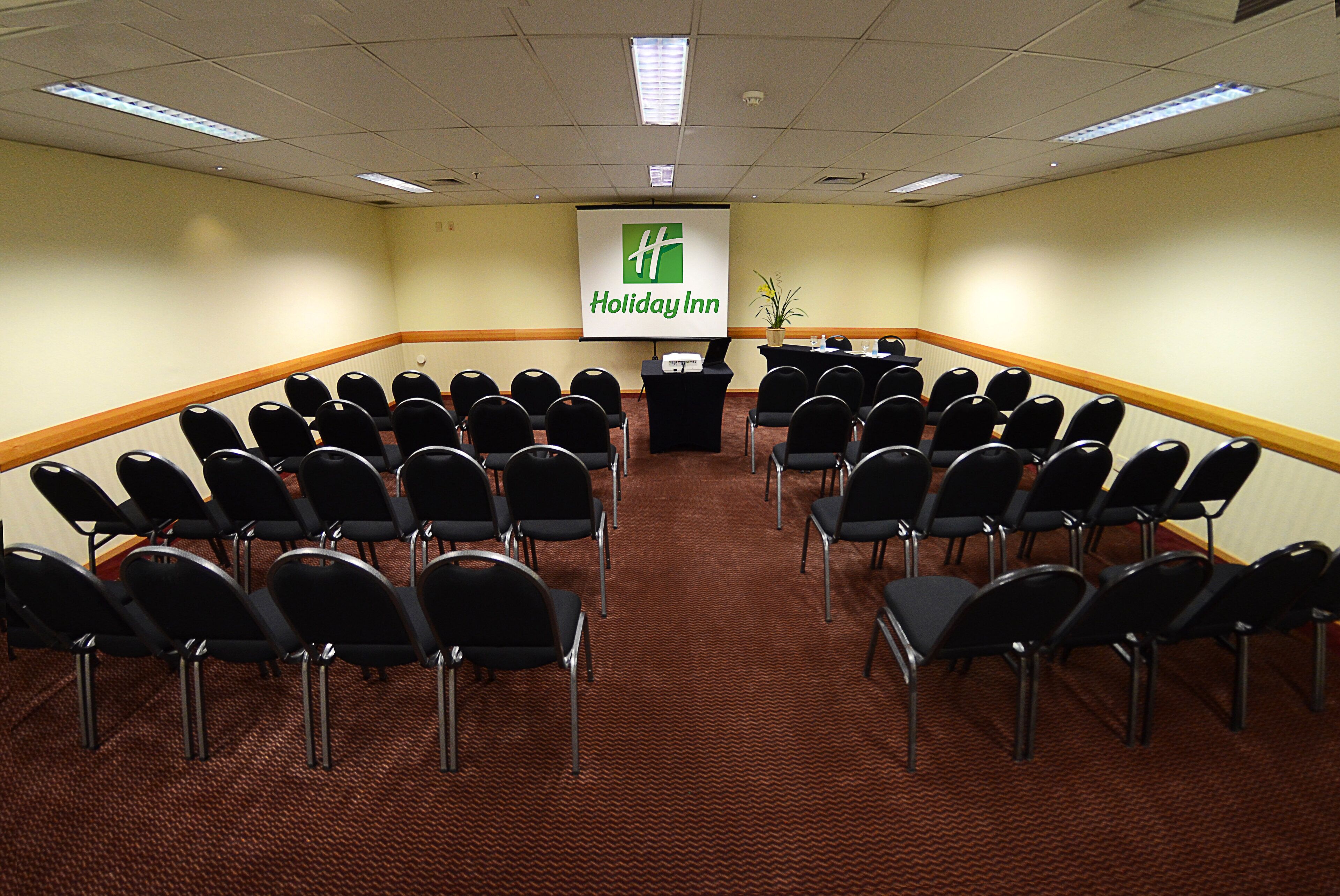 Uma sala de conferência com fileiras de cadeiras pretas, um púlpito e uma tela exibindo o logotipo do Holiday Inn.