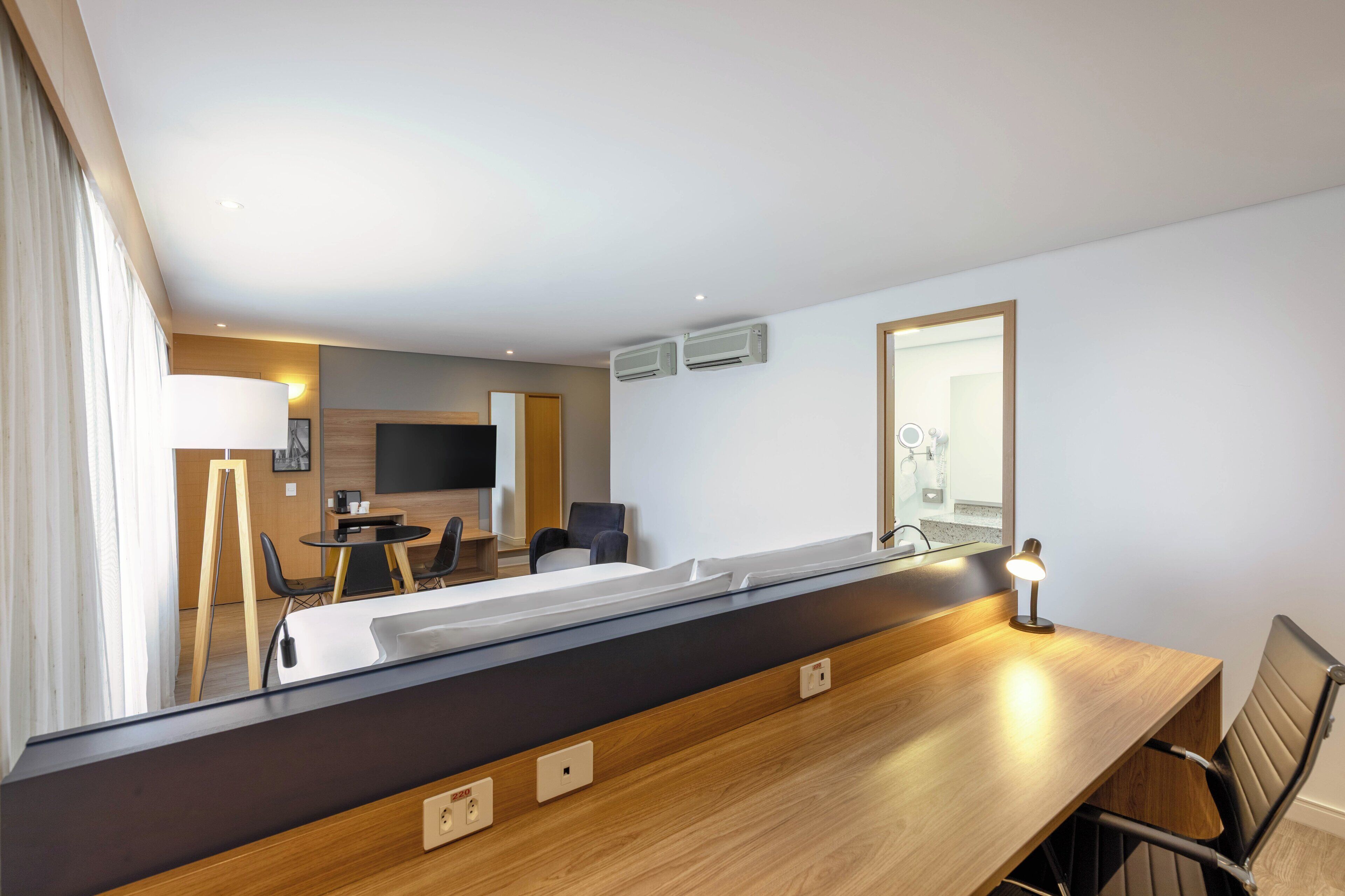 Quarto de hotel moderno com uma mesa de madeira comprida, um espelho grande e uma televisão de tela plana.
