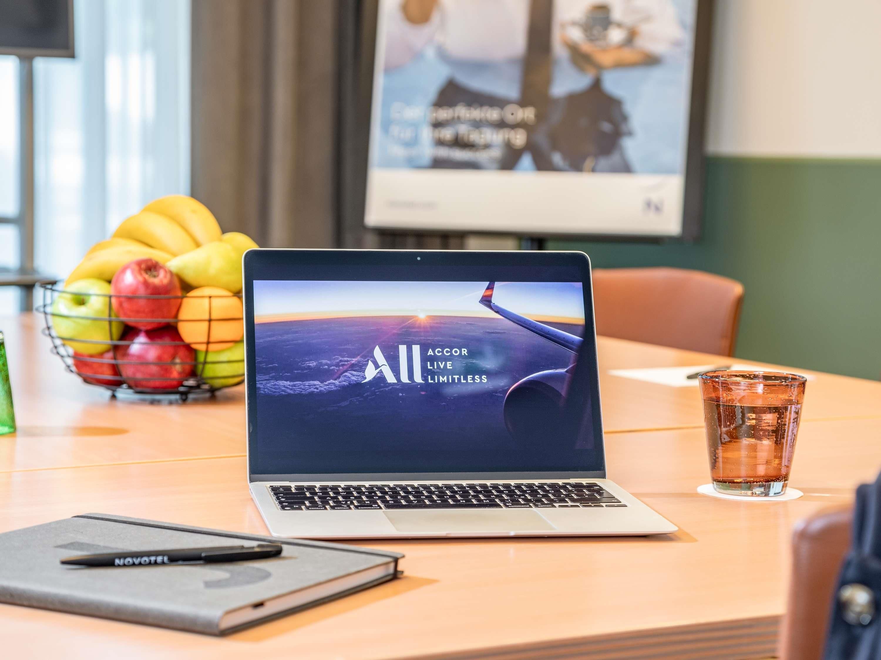 Um laptop sobre uma mesa com uma fruteira, um copo de água e um caderno.