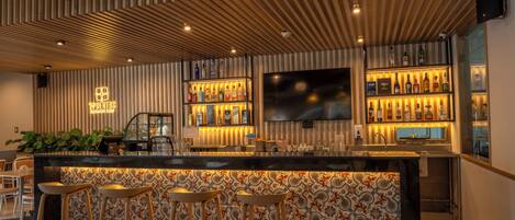 Uma área de bar moderna, com teto de ripas de madeira, balcão e banquetas, e uma TV fixada na parede.