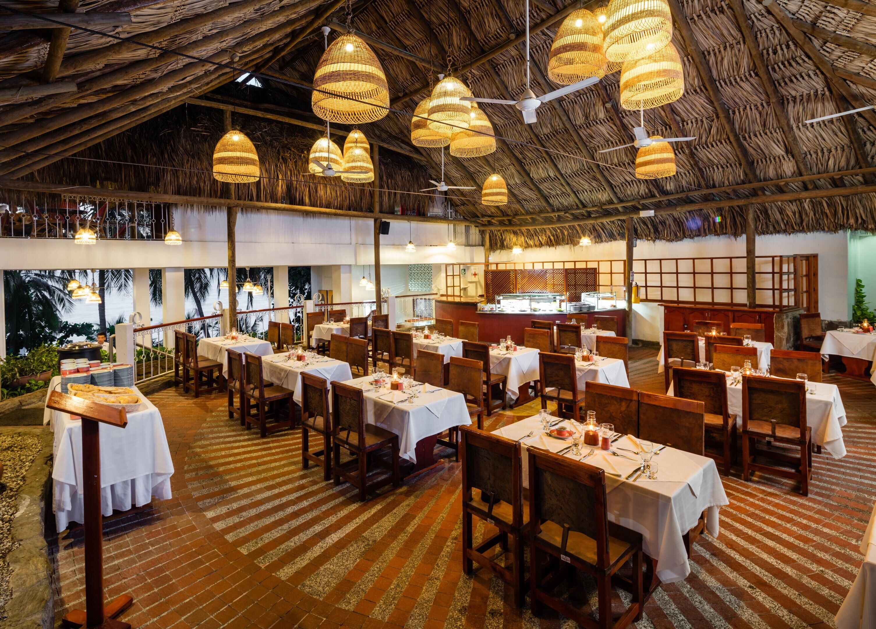 Um restaurante com várias mesas e cadeiras, um telhado de palha e luminárias de vime penduradas.