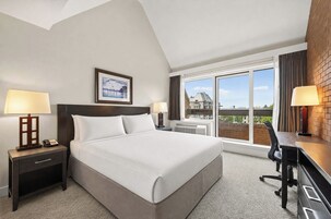 Quarto de hotel com uma cama grande, duas mesas de cabeceira com luminárias, uma escrivaninha com cadeira e uma janela ampla com vista para edifícios.