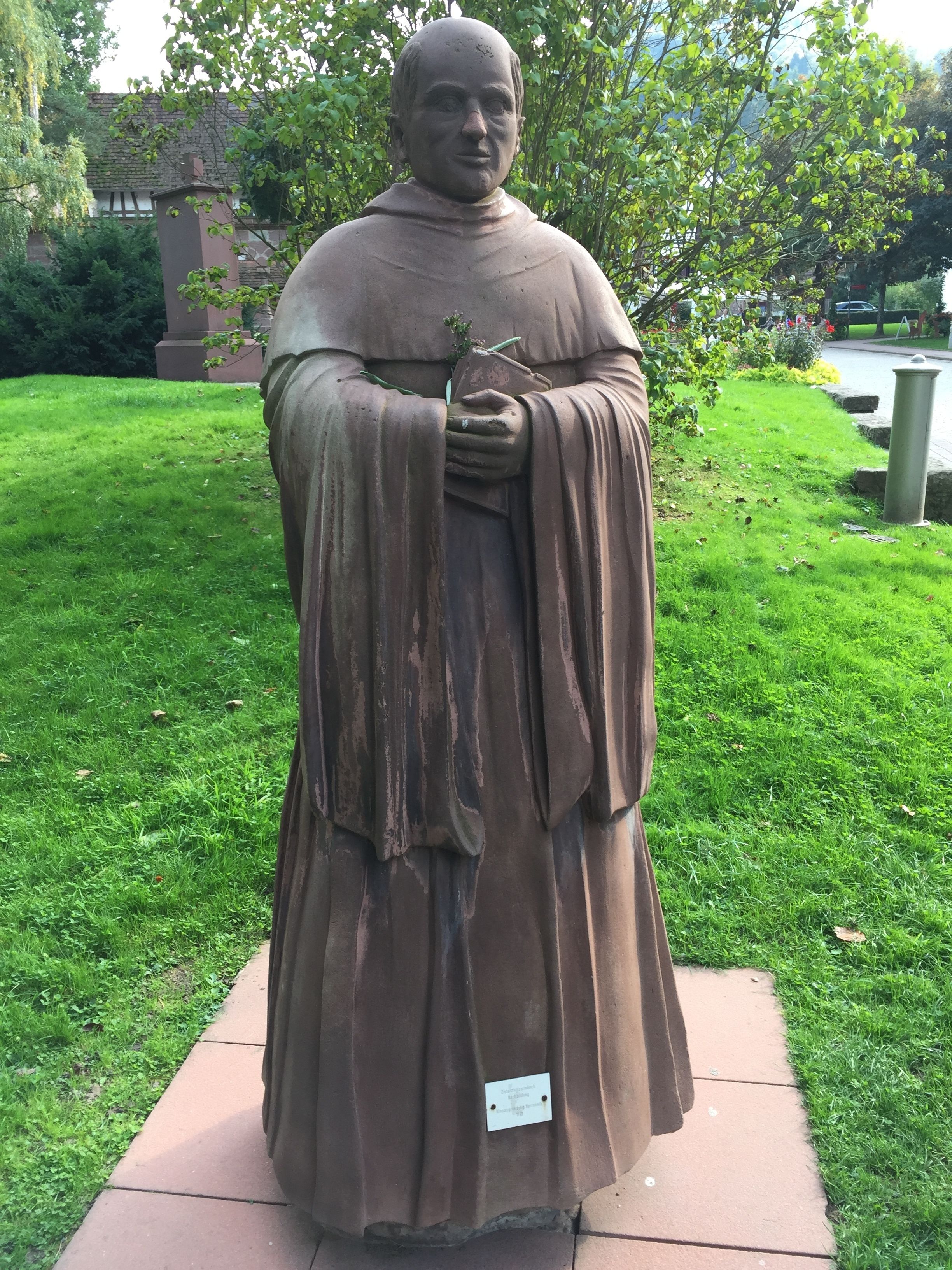 Uma estátua de uma figura vestida com manto, segurando um livro, sobre uma área pavimentada, com grama e árvores ao fundo.