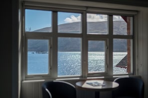 Quarto com uma janela grande com vista para um lago e montanhas. Há uma pequena mesa com cadeiras em primeiro plano.