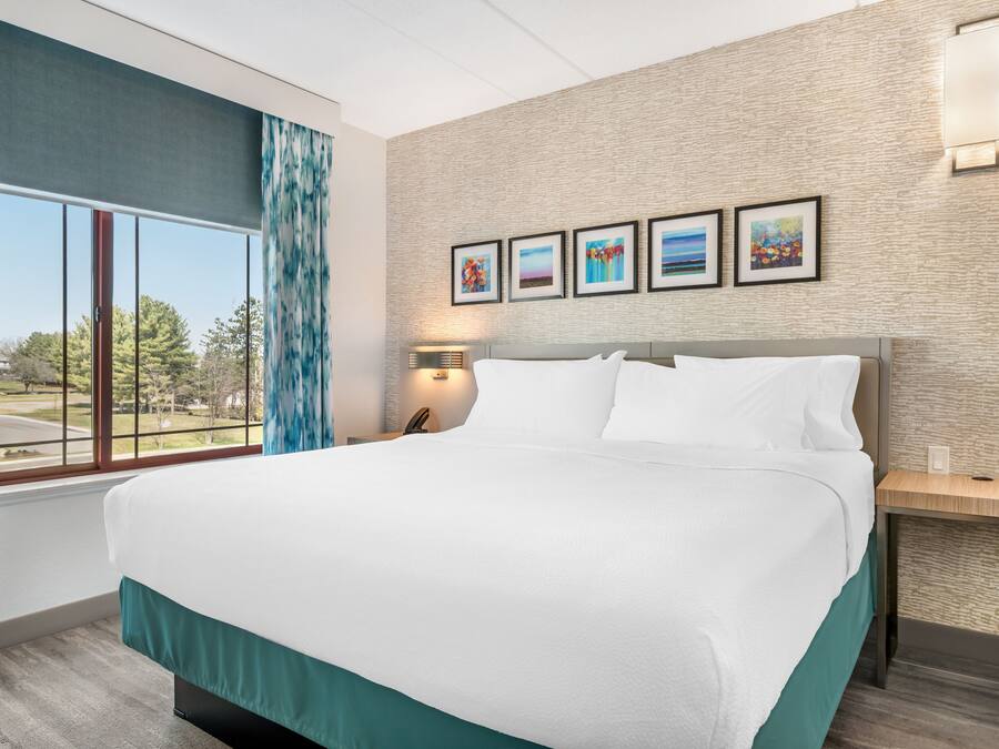 Quarto de hotel com uma cama grande, uma janela com cortinas, uma parede com quadros emoldurados e uma mesa de cabeceira com abajur.