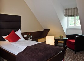 Quarto de hotel com cama, mesa de cabeceira com abajur, uma cadeira e pequena mesa com frutas.