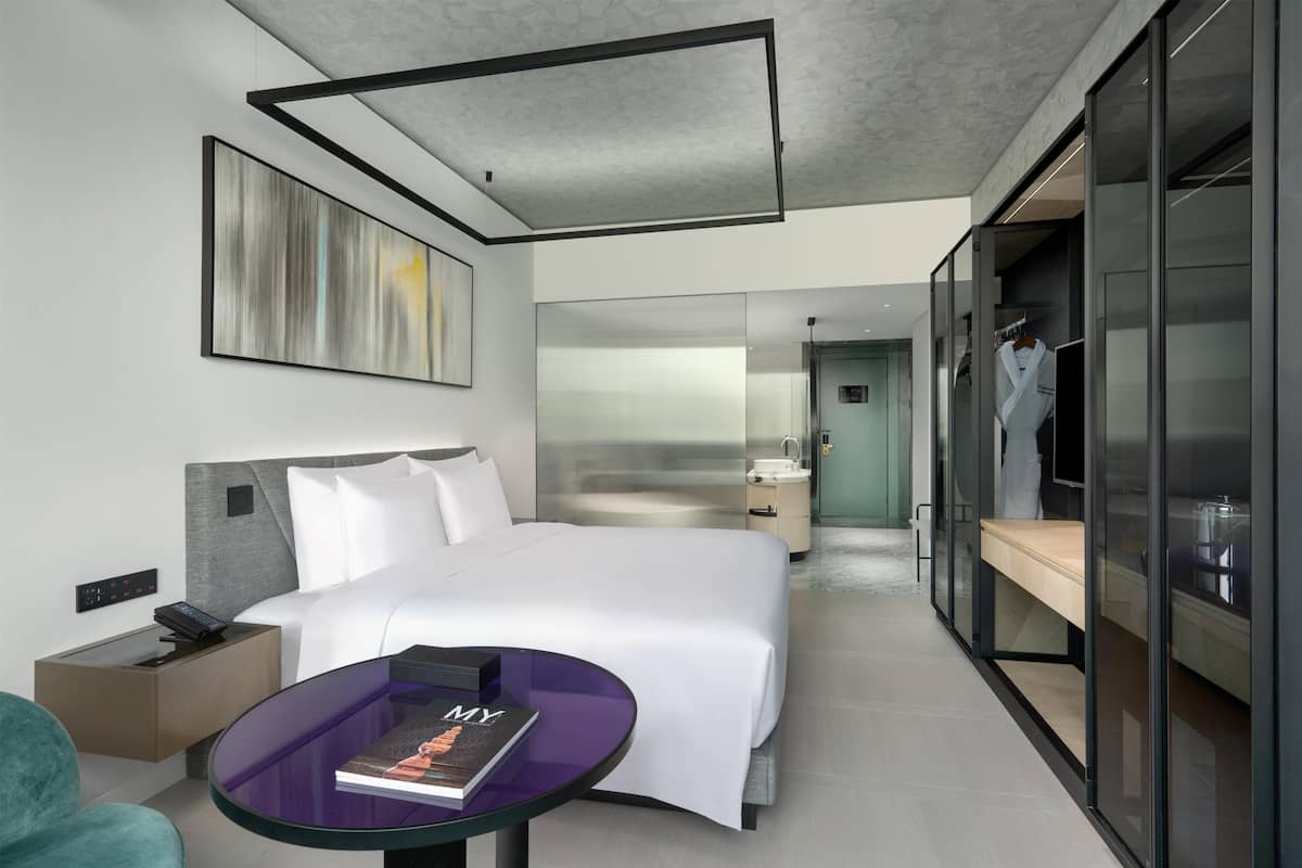 Quarto de hotel moderno com uma cama grande, uma mesa de vidro com um livro, uma mesinha de cabeceira, um guarda-roupa e um banheiro visível através de uma divisória de vidro.