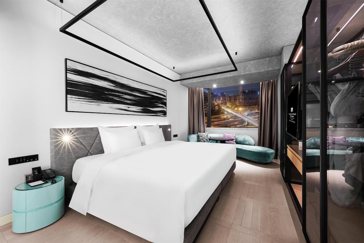 Quarto de hotel moderno com uma cama grande, mesa de cabeceira com telefone, divisória de vidro e vista para a cidade.