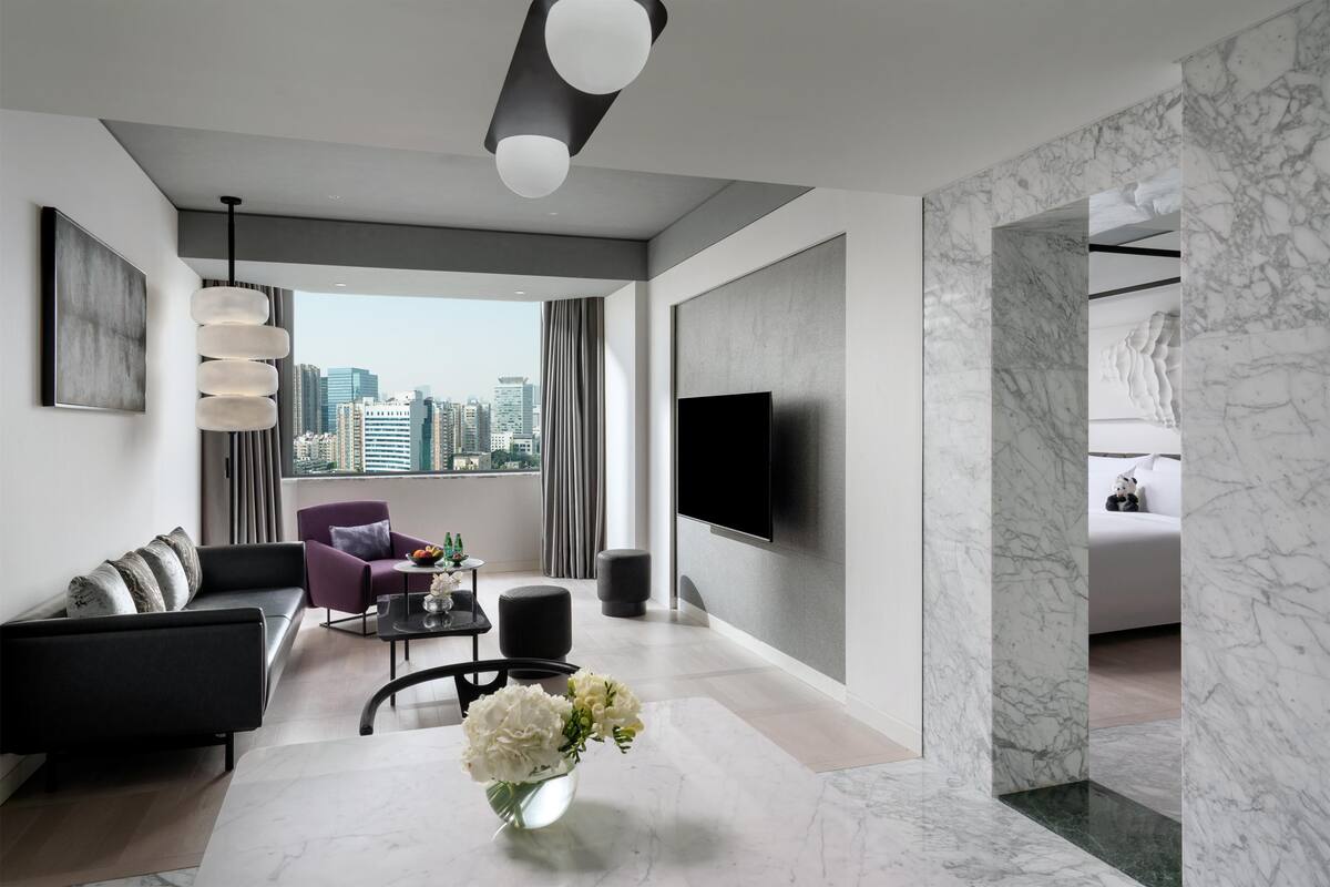 Sala moderna com sofá, poltrona, mesa de centro e um vaso de flores. Uma TV de tela plana está montada na parede, e uma vista da cidade é visível pela janela.