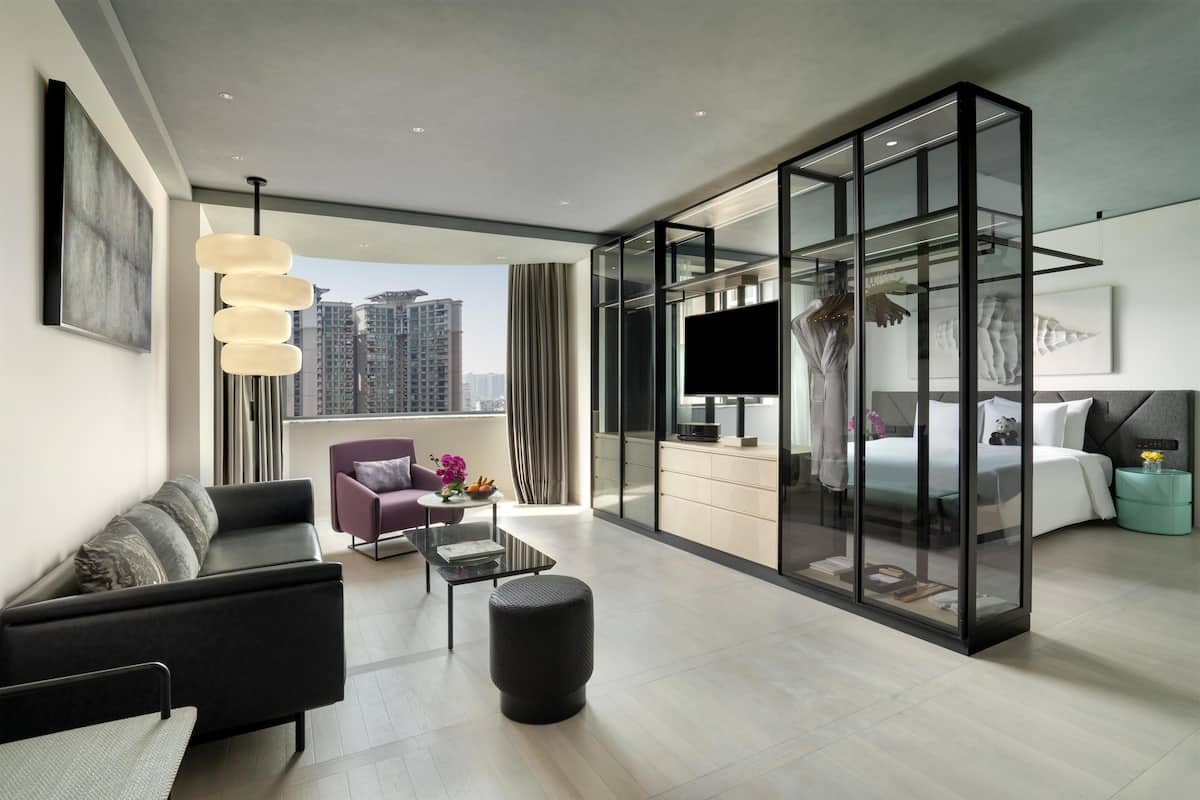Quarto de hotel moderno com sofá, mesa de centro, televisão, cama e divisória de vidro.