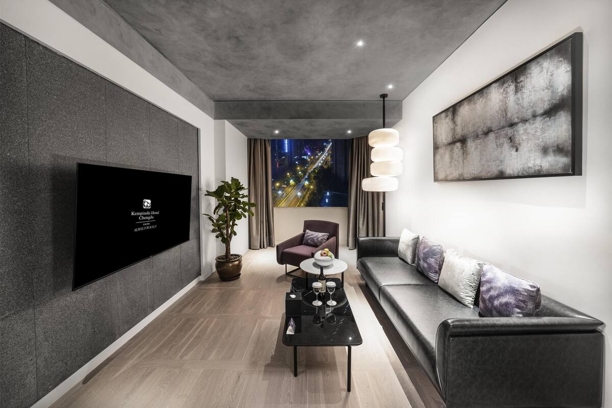 Uma sala de estar moderna com televisão de tela plana, uma mesa de centro com copos de vinho, um sofá e um grande quadro abstrato.