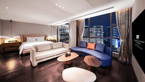 Quarto de hotel moderno com cama, sofá e duas mesas de centro. O quarto tem vista para a cidade através de grandes janelas.