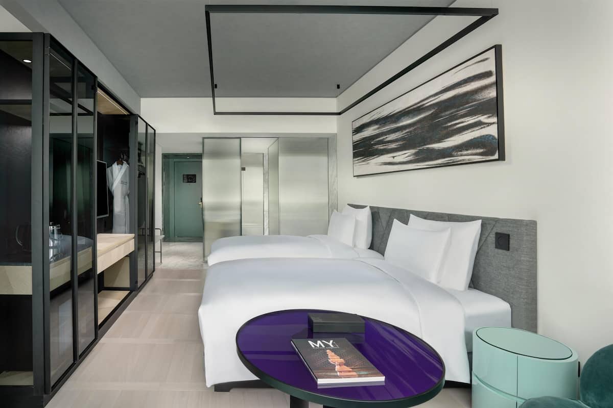 Quarto de hotel moderno com uma cama grande, uma mesa de vidro com um livro, uma lixeira verde e uma grande pintura abstrata na parede.