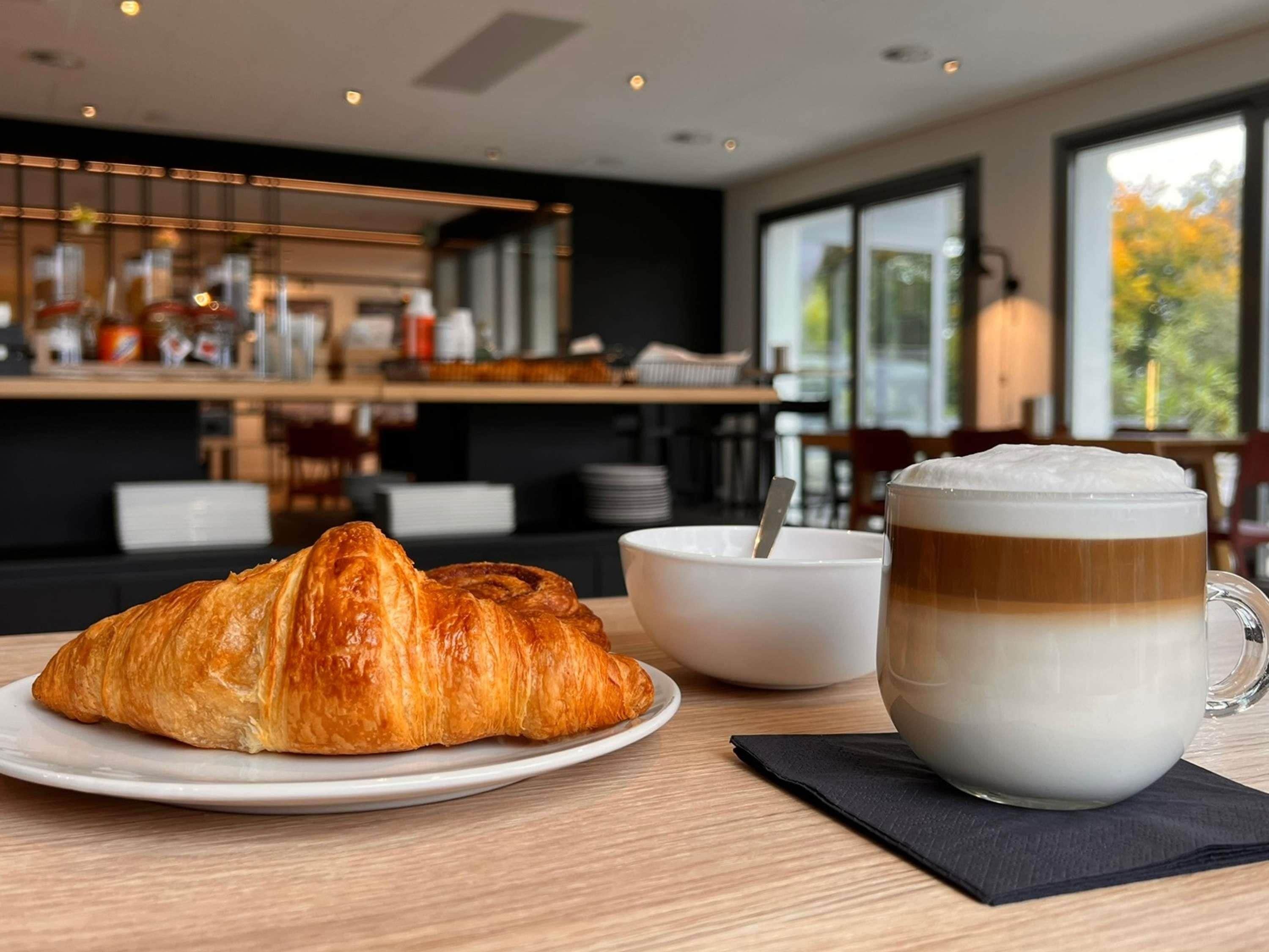 Um croissant num prato, uma tigela com uma colher e um copo de café sobre uma mesa em um café moderno.