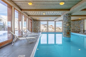 Piscina interior com paredes de pedra e vigas de madeira, rodeada por espreguiçadeiras e grandes janelas com vista para uma paisagem nevada.