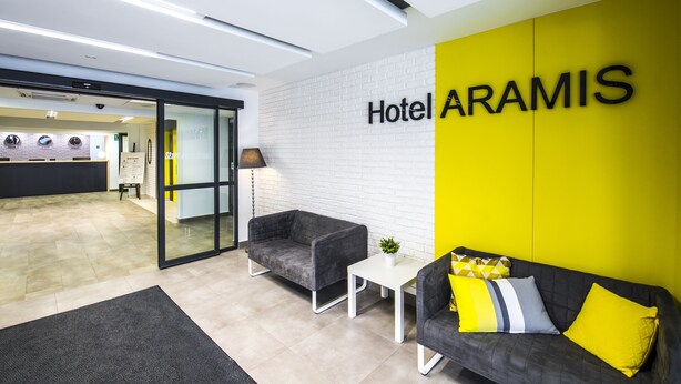 Uma área de recepção de hotel com design moderno, com balcão de recepção, sala de estar com dois sofás, uma pequena mesa com luminária e uma parede de destaque amarelo vibrante com o nome do hotel.