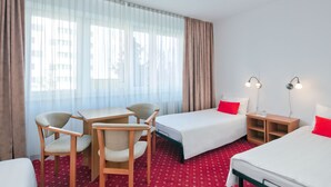 Quarto de hotel com duas camas de solteiro, uma escrivaninha com cadeiras, uma mesinha e uma mesa de cabeceira com abajures.