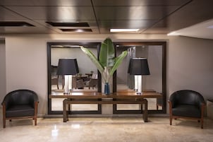 Um lobby com um banco de madeira, duas poltronas de couro preto, um espelho grande e um vaso com planta.