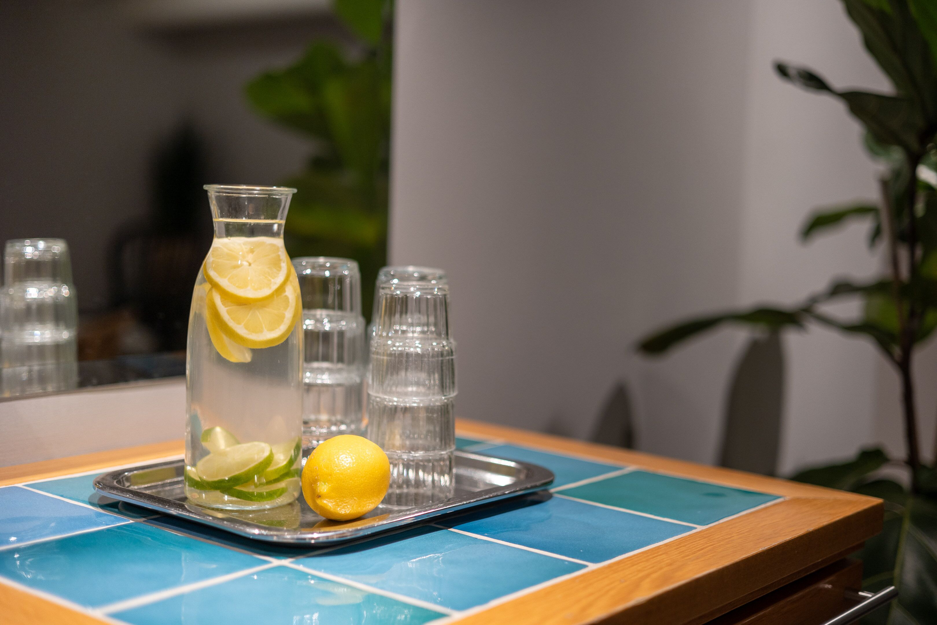 Um jarro de vidro transparente com rodelas de limão e um limão numa bandeja, rodeado por copos de vidro numa superfície de azulejos.