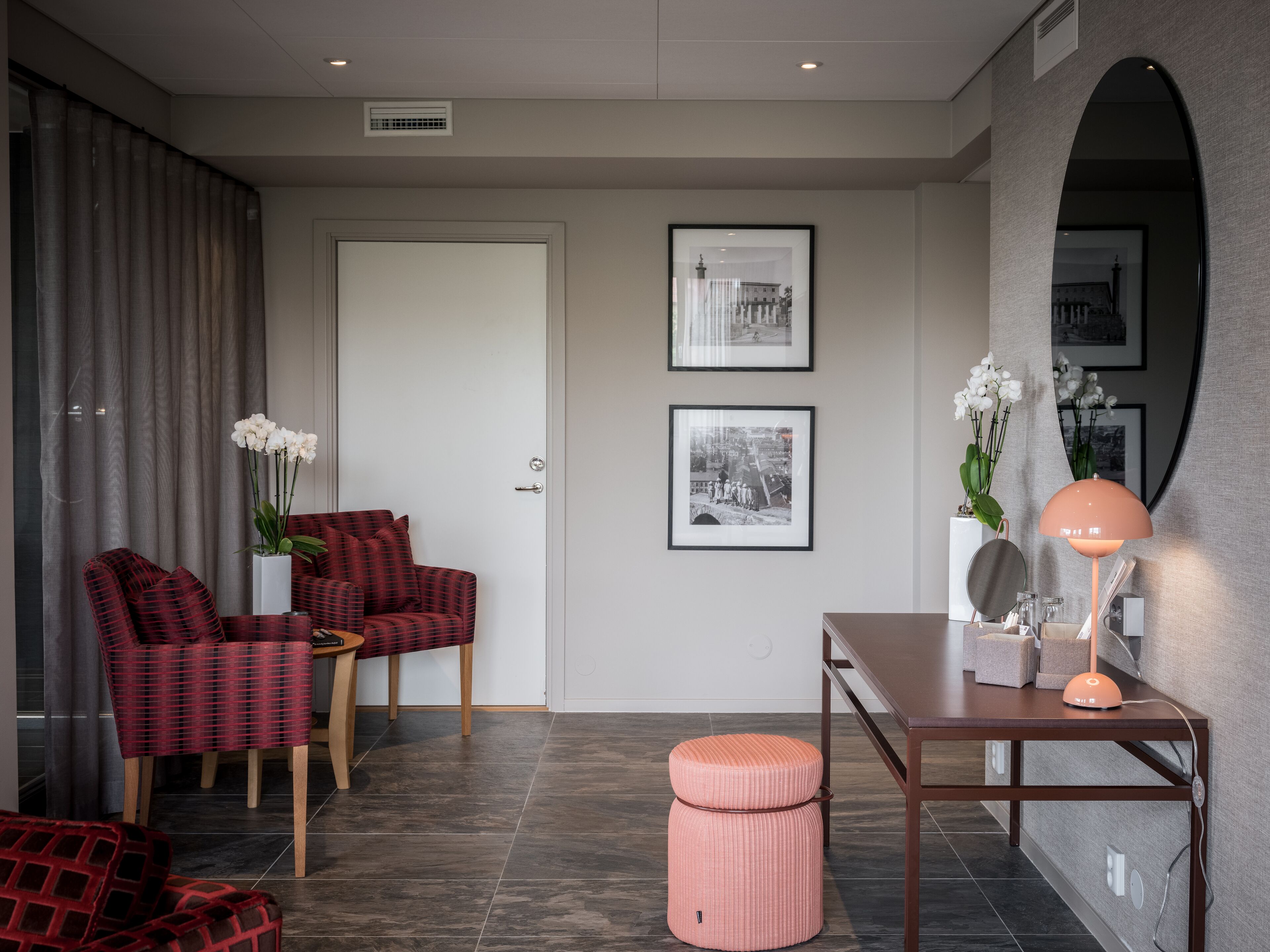 Uma sala de estar moderna com um sofá xadrez vermelho, uma mesa de centro de madeira, um banquinho rosa e uma escrivaninha com uma luminária. Há duas fotografias em preto e branco emolduradas na parede.